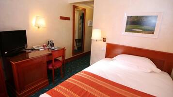 Quarto de hotel com cama, escrivaninha com televisão, cadeira, abajur e um quadro na parede.