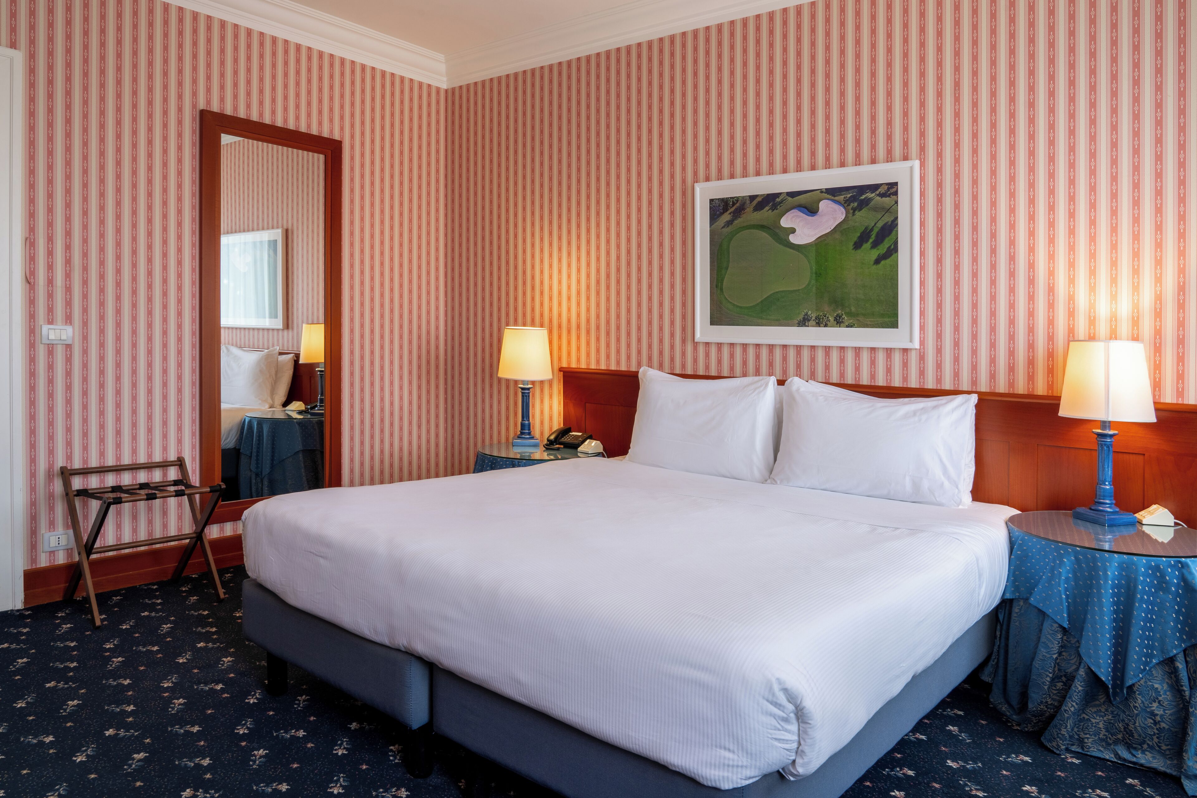 Quarto de hotel com uma cama grande, duas mesas de cabeceira com abajures, uma cadeira e um quadro na parede.