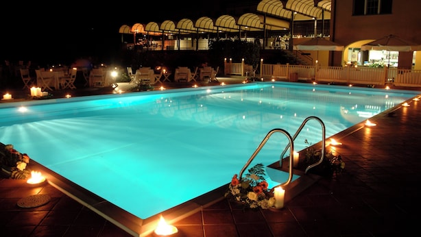 Piscina com deck revestido, rodeada por espreguiçadeiras e guarda-sóis, e um edifício com terraço coberto ao fundo.