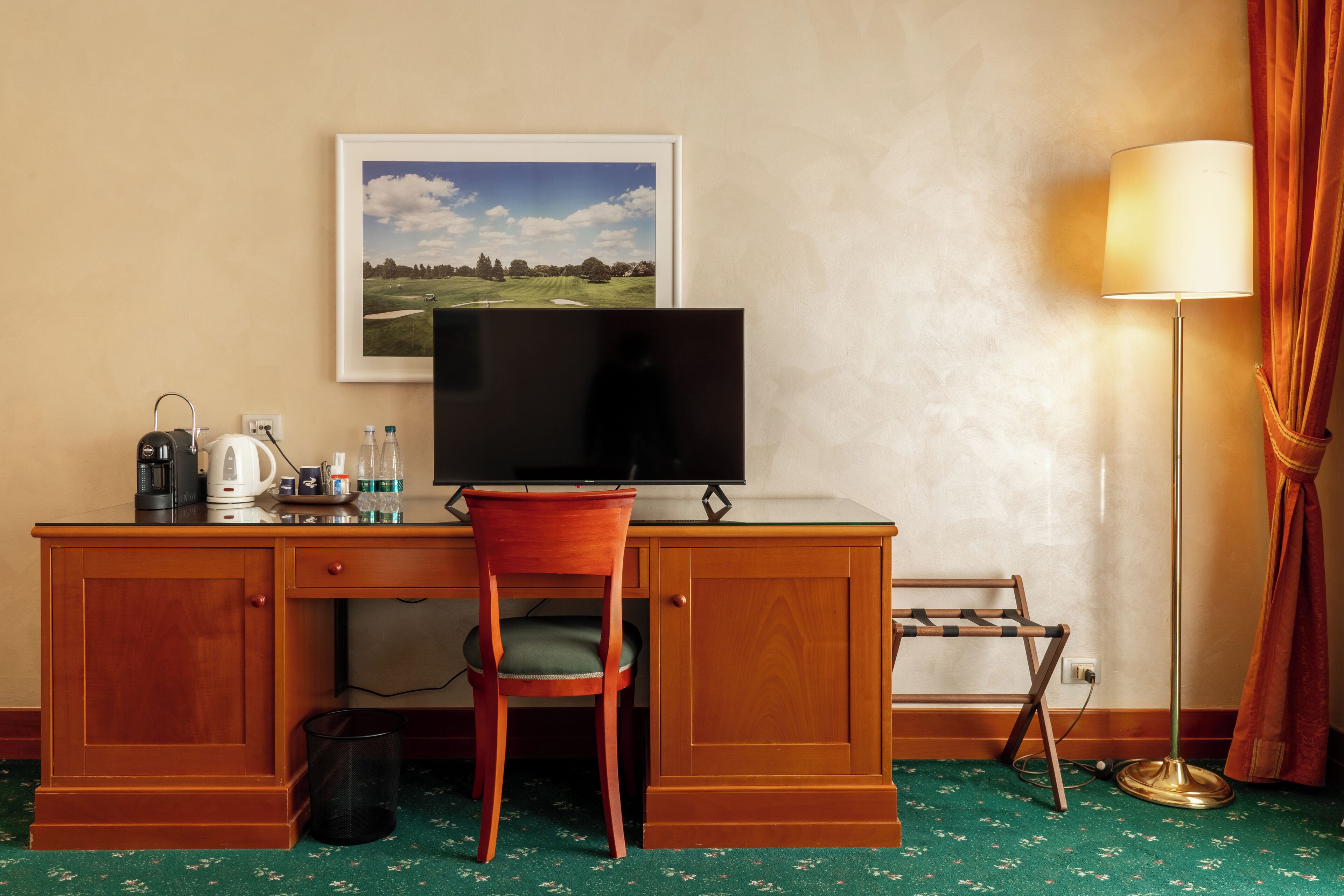 Quarto de hotel com escrivaninha de madeira, televisão, quadro decorativo de paisagem, luminária de chão e janela com cortinas.