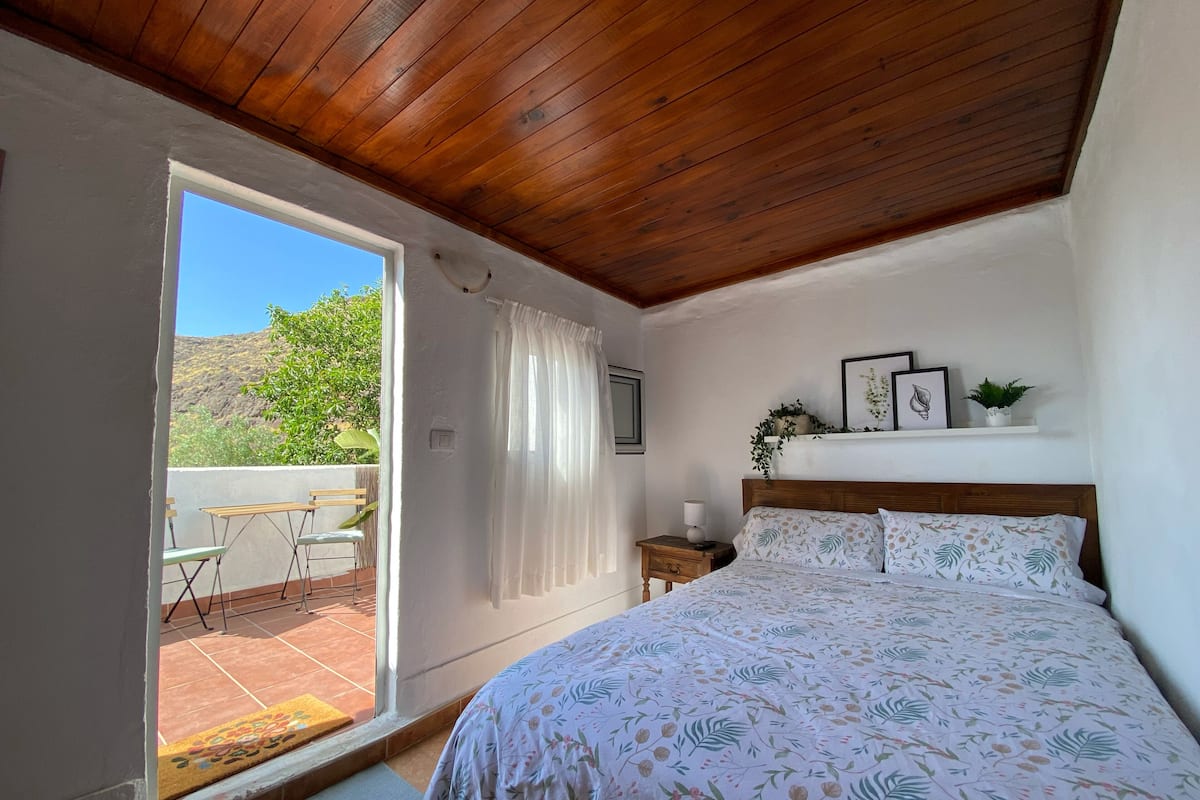 Quarto com teto de madeira, cama com roupa de cama florida, mesa de cabeceira, prateleira com plantas e fotos, e porta de vidro deslizante que dá para um pátio externo.