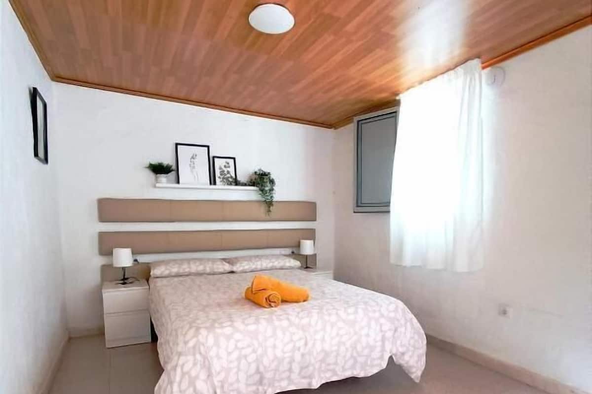 Quarto com teto de madeira, cama com colcha estampada em branco, mesa de cabeceira e prateleira na parede com objetos decorativos.