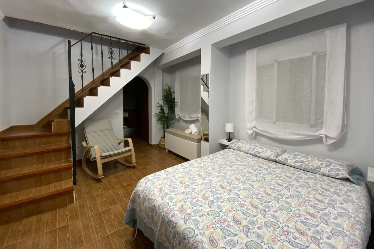 Quarto com escada de madeira, cama com roupa de cama estampada, mesa de cabeceira com abajur e janela com cortinas.