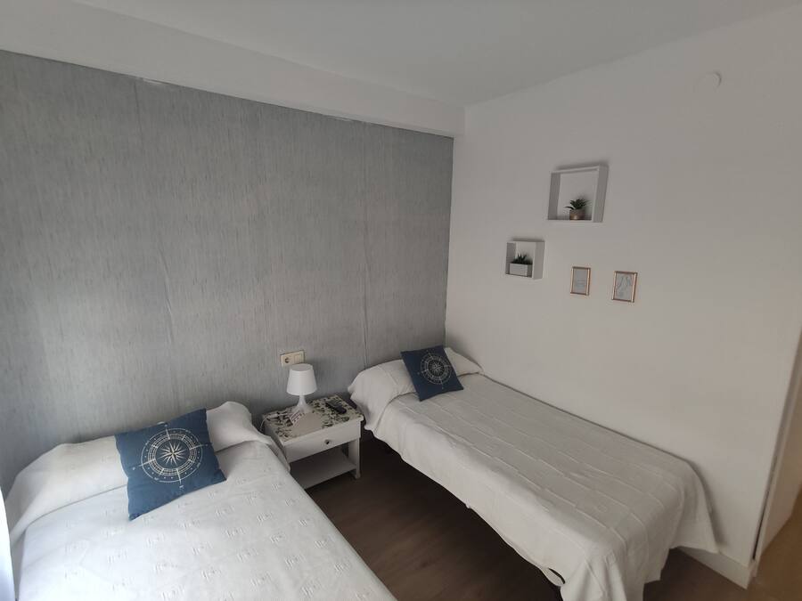 Quarto com duas camas de solteiro, mesa de cabeceira, luminária e uma pequena prateleira com uma planta.
