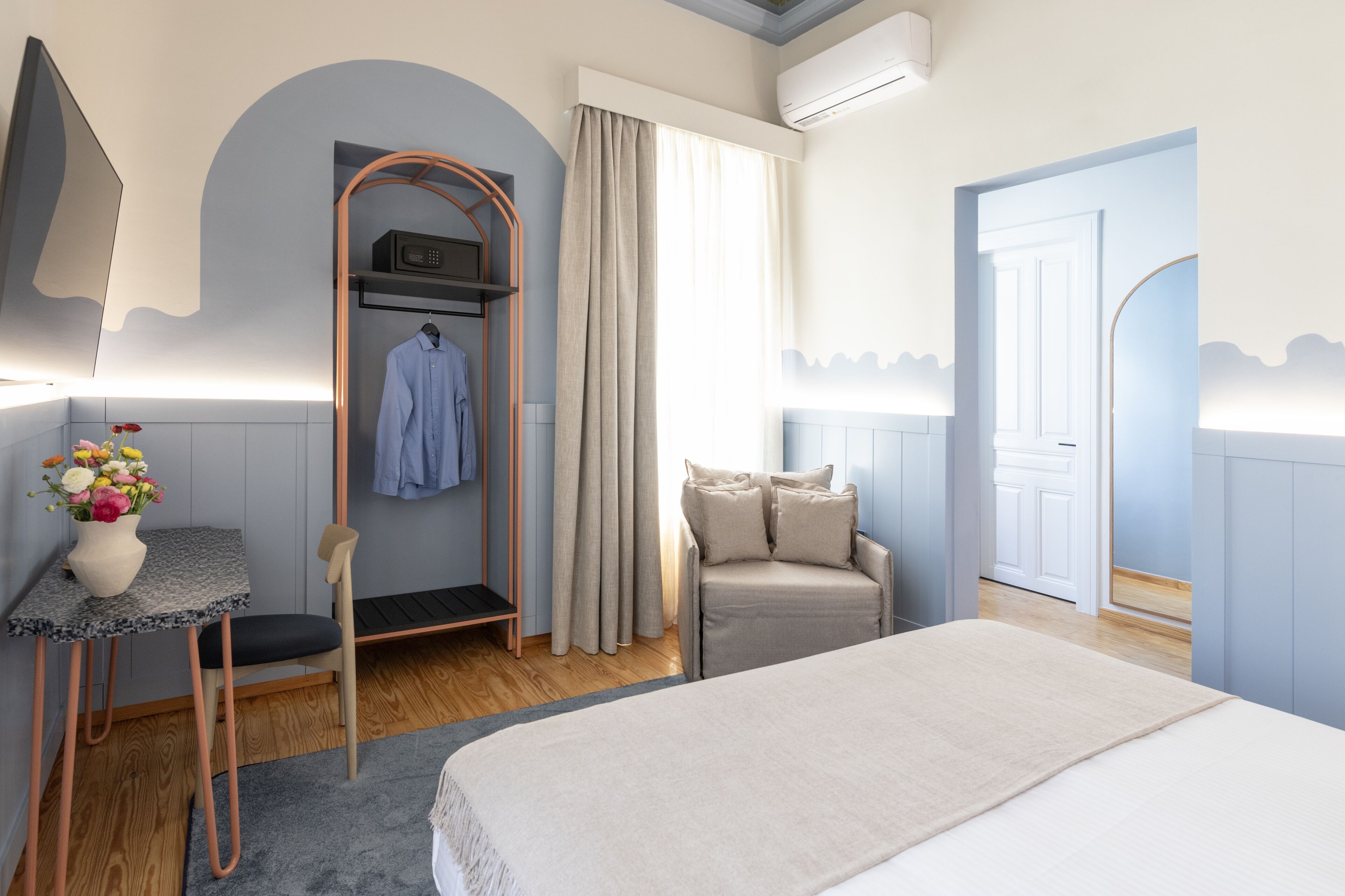 Quarto de hotel com cama, cadeira, mesa com um vaso de flores, cabideiro e janela com cortinas.