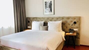 Quarto de hotel com cama grande, cabeceira estofada, mesa de cabeceira com abajur e quadro na parede.