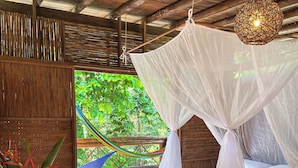 Uma cama de madeira com dossel branco, luminária pendente e vista para o verde lá fora.