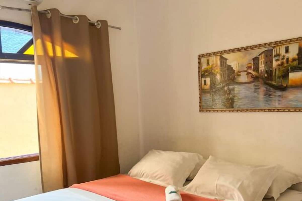Uma cama de solteiro com lençóis brancos e um cobertor cor de pêssego, um quadro com uma cena de canal na parede e uma janela com cortina.