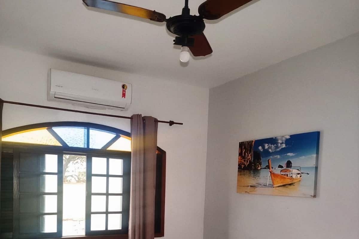 Quarto de hotel com cama, ventoinha no teto, janela com cortinas, aparelho de ar condicionado e quadro com cena de praia.