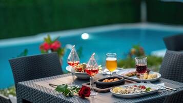 Mesa de jantar com pratos de comida, copos de vinho e um recipiente com petiscos ao lado de uma piscina.