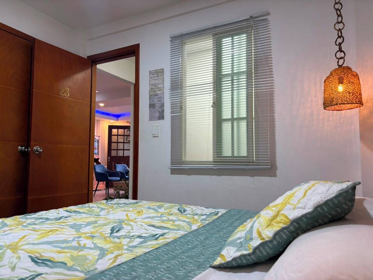 Um quarto com cama, janela com persianas, luminária pendente e porta.