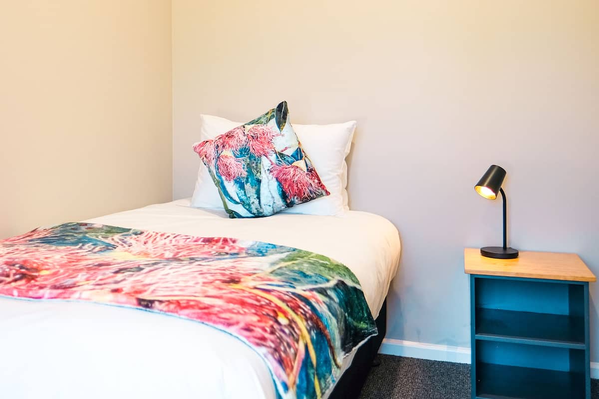 Uma cama bem arrumada com um cobertor colorido e um travesseiro decorativo, uma mesa de cabeceira com uma luminária e uma estante azul.