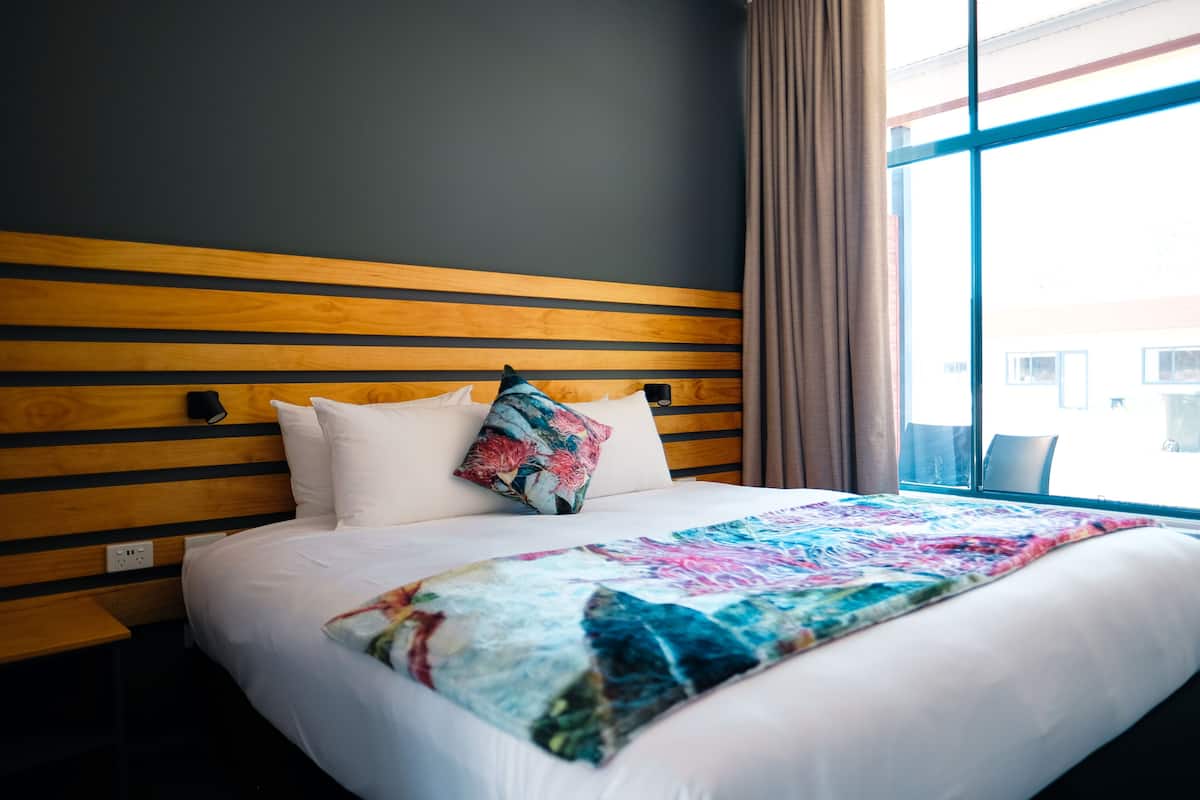 Quarto de hotel com cabeceira de madeira, roupa de cama branca, colcha colorida e uma grande janela com cortinas.
