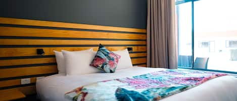 Quarto de hotel com cabeceira de madeira, roupa de cama branca, colcha colorida e uma grande janela com cortinas.