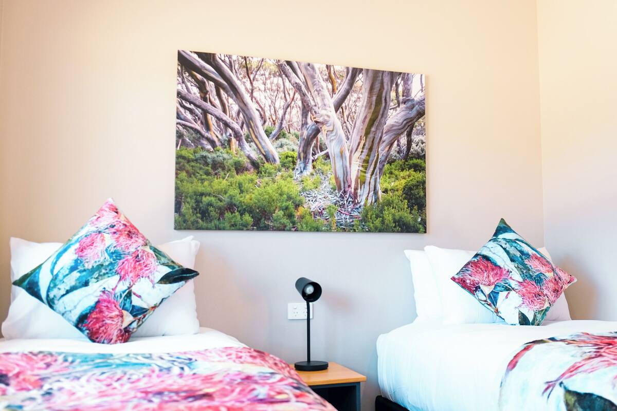 Quarto de hotel com duas camas, cada uma com colcha estampada de flores, uma mesa de cabeceira com abajur e um quadro de árvores fixado na parede.