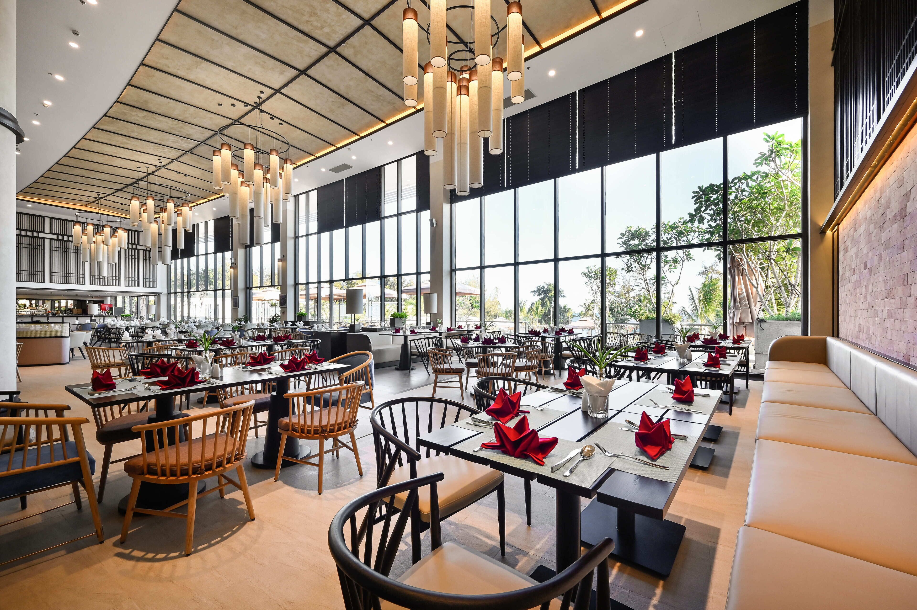 Um restaurante espaçoso com pé-direito alto, grandes janelas e luminárias modernas. As mesas estão bem dispostas com cadeiras, e o interior combina elementos de madeira e metal.