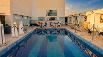 Uma área de piscina com piso em azulejo, um bar com banquetas e um espaço de estar com cadeiras.