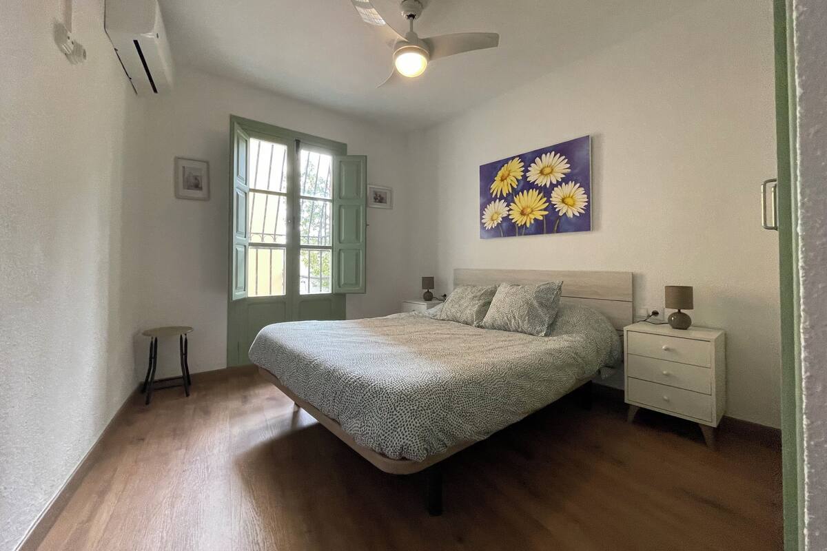 Quarto com cama, mesa de cabeceira, ventoinha no teto e janela com persianas.