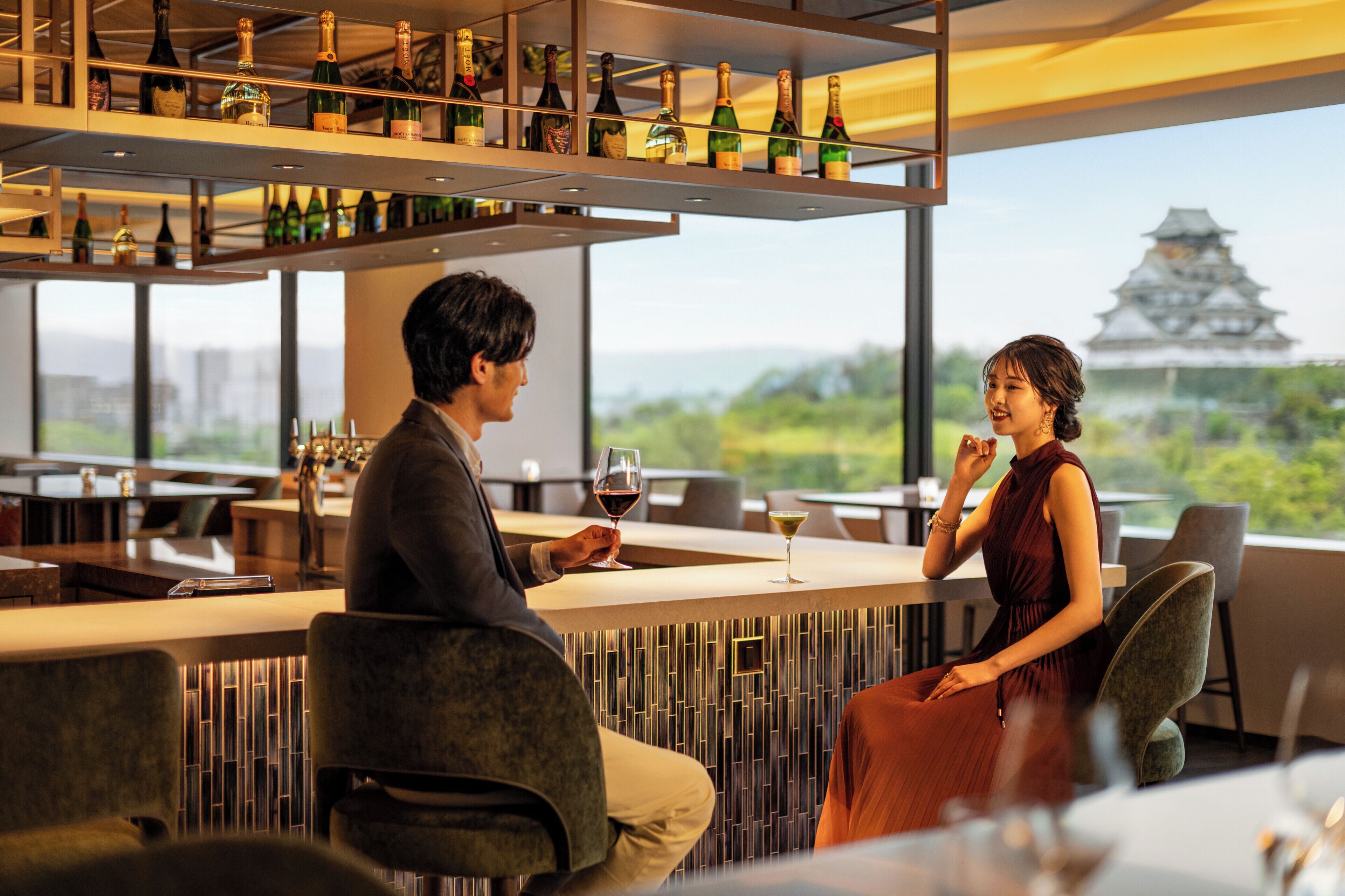 Uma área de bar moderna com diversos frascos nas prateleiras, um casal sentado à mesa com vista para um castelo japonês tradicional ao fundo.