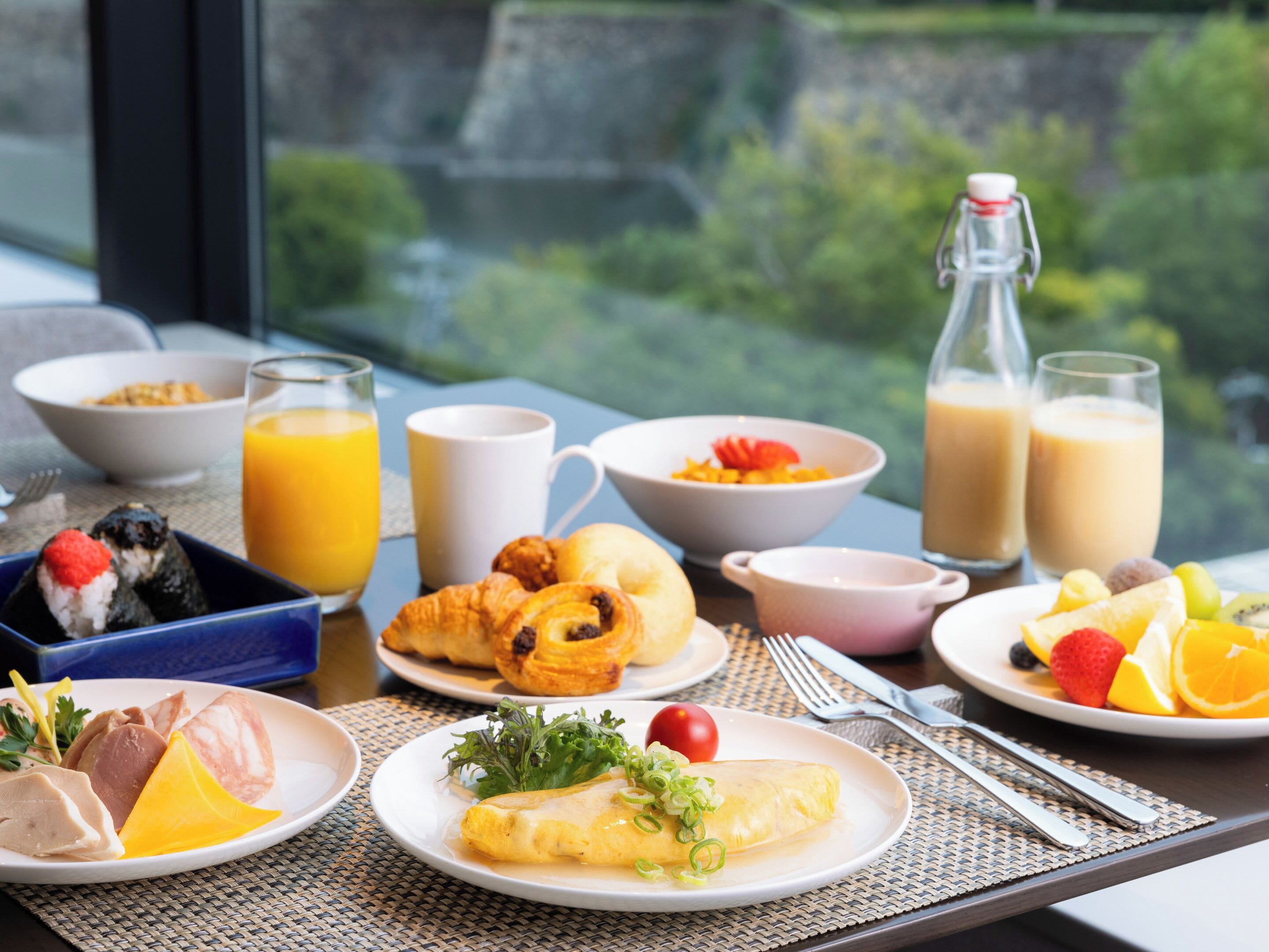 Mesa de café da manhã com diversos pratos, incluindo croissants, frutas e uma garrafa de suco, com vista para o verde.