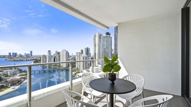 Uma varanda com cadeiras brancas, uma pequena mesa e um vaso de planta. Uma linha do horizonte urbana e a orla marítima são visíveis ao fundo.
