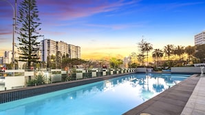 Uma piscina com borda azulejada, cercada por uma grade, e palmeiras ao fundo.