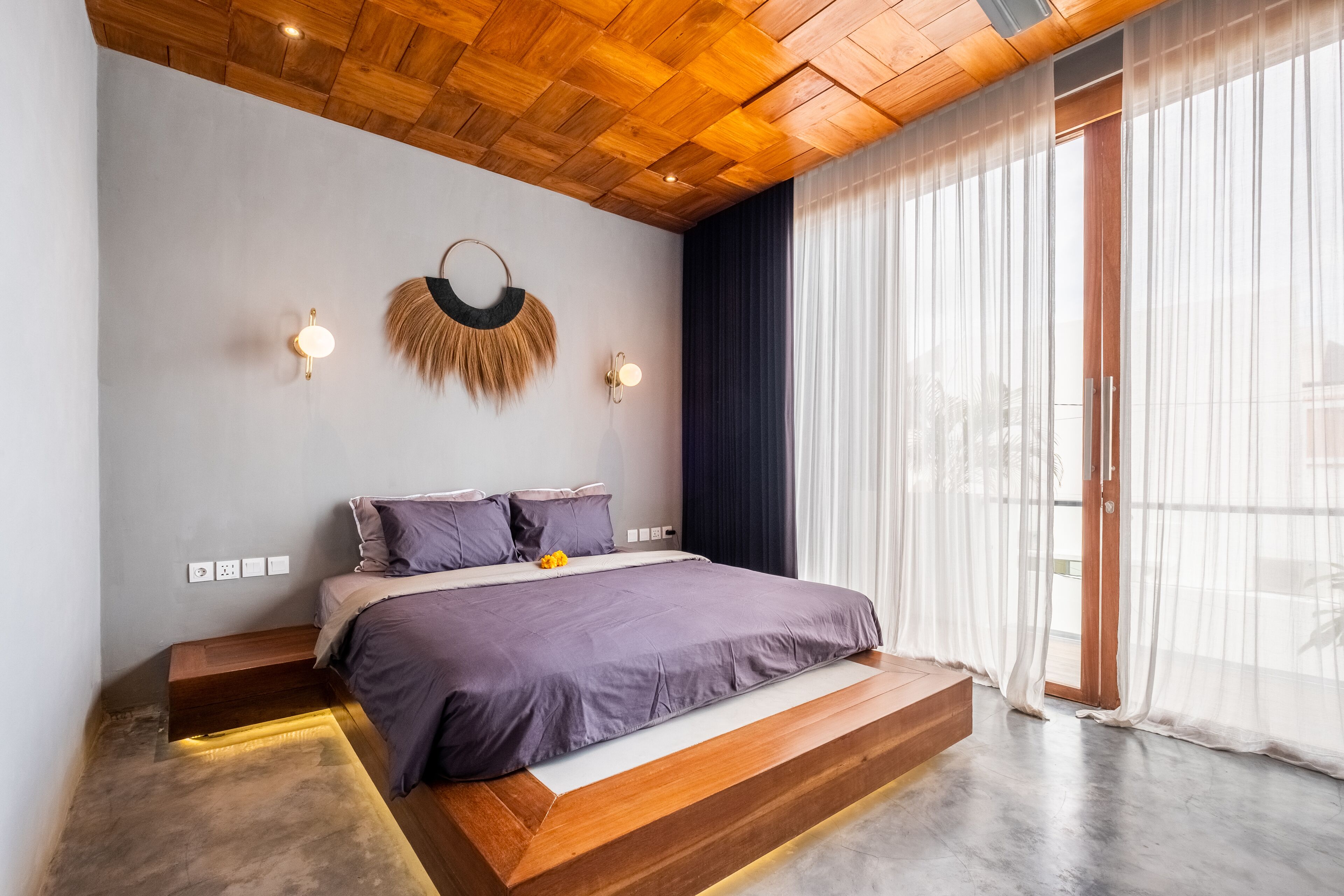 Quarto moderno com teto de madeira, cama com estrutura de madeira e uma janela grande com cortinas leves.