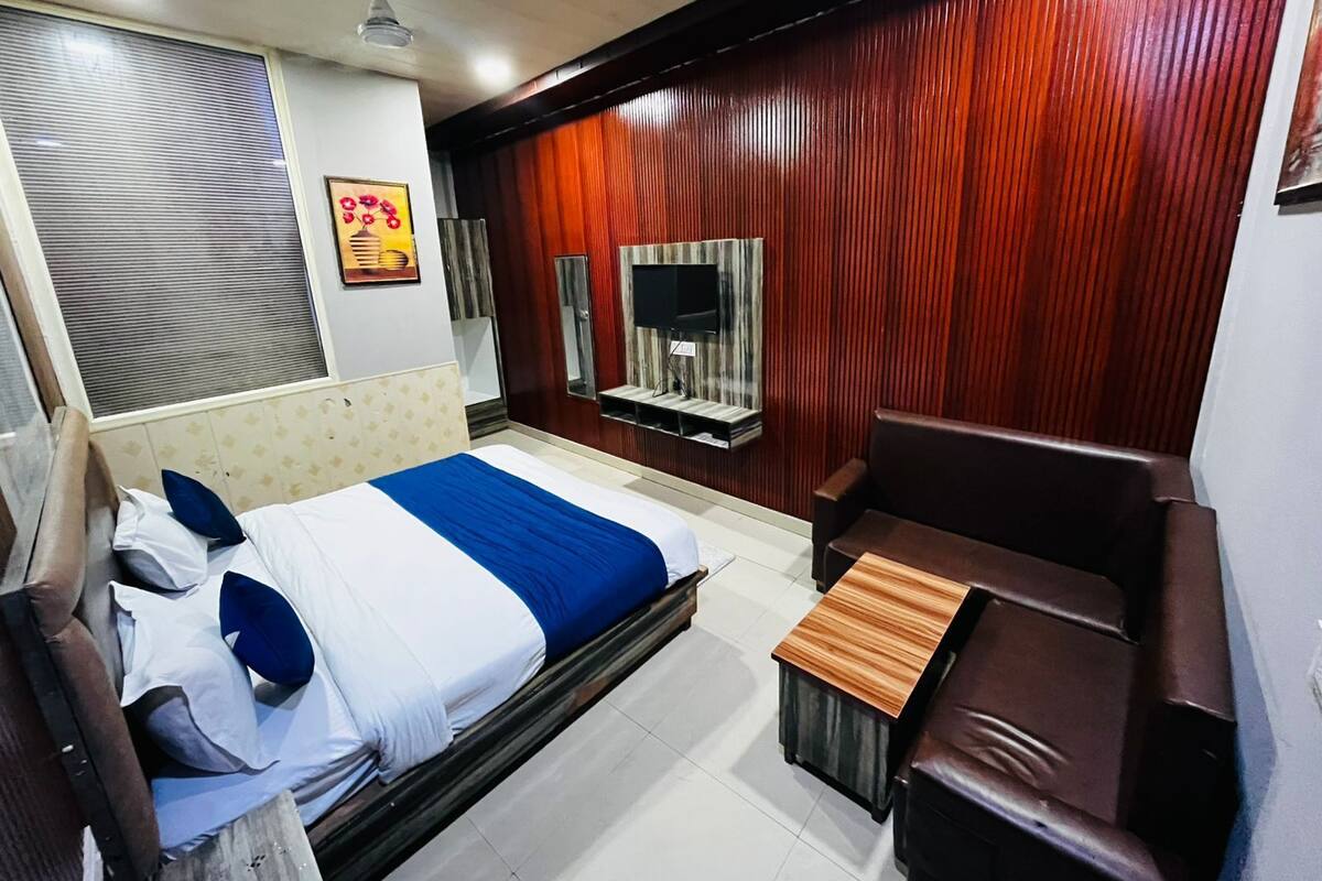 Quarto de hotel com cama, uma pequena mesa de madeira, televisão fixada na parede e janela com persianas.