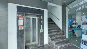 Entrada de um edifício moderno, com porta de vidro, escada e sinalização indicando área de vacinação e higienização.