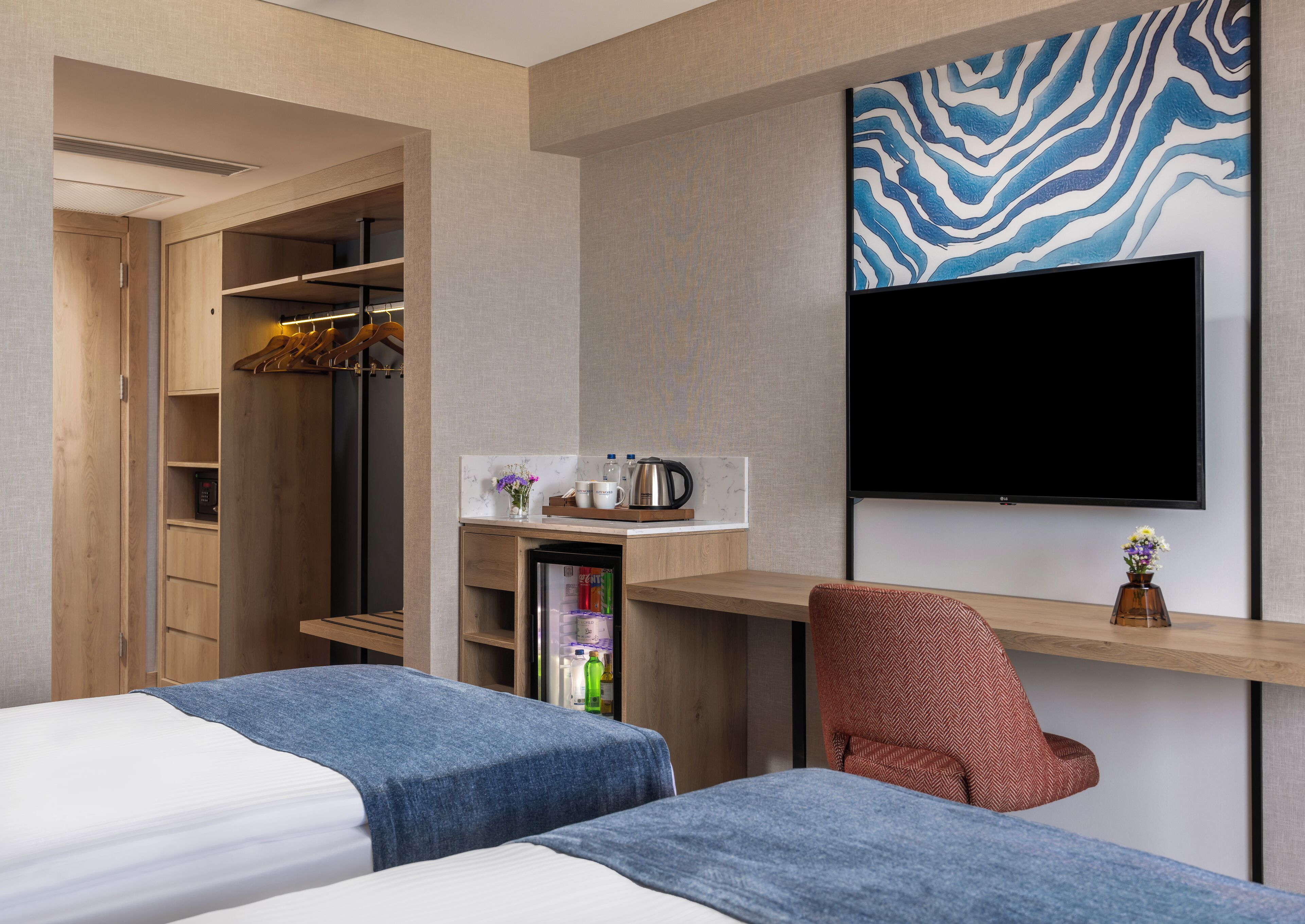 Quarto de hotel com duas camas, televisão, guarda-roupa, pequena cozinha e uma parede com um quadro abstrato azul.
