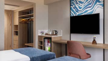 Quarto de hotel com duas camas, televisão, guarda-roupa, pequena cozinha e uma parede com um quadro abstrato azul.