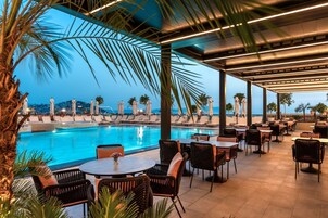 Uma área à beira da piscina com espreguiçadeiras e mesas, uma piscina e vista para a cidade.