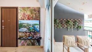 Um saguão de hotel com porta de madeira, uma televisão grande fixada na parede, uma área de estar com almofadas de macramé e uma parede decorativa com plantas.