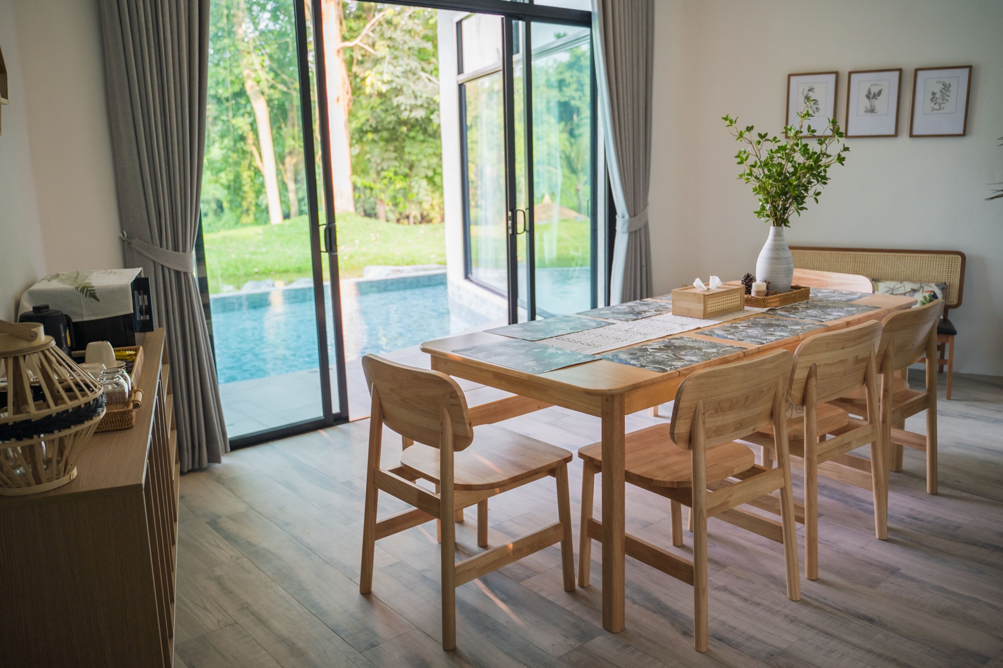 Uma sala de jantar com mesa e cadeiras de madeira, um vaso com planta e vista para uma piscina através de grandes portas de vidro.