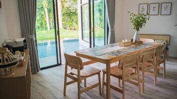 Uma sala de jantar com mesa e cadeiras de madeira, um vaso com planta e vista para uma piscina através de grandes portas de vidro.