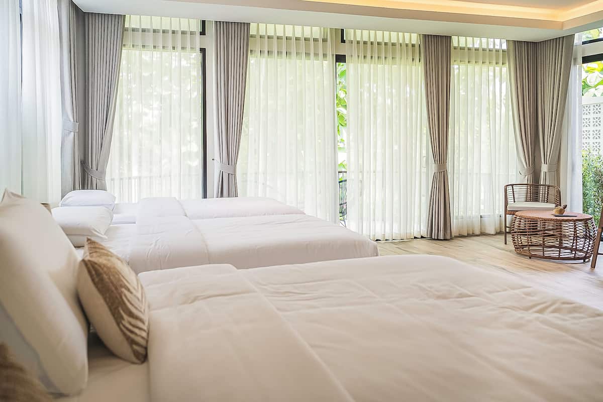 Quarto de hotel com duas camas grandes, uma mesa redonda pequena e uma cadeira de vime.