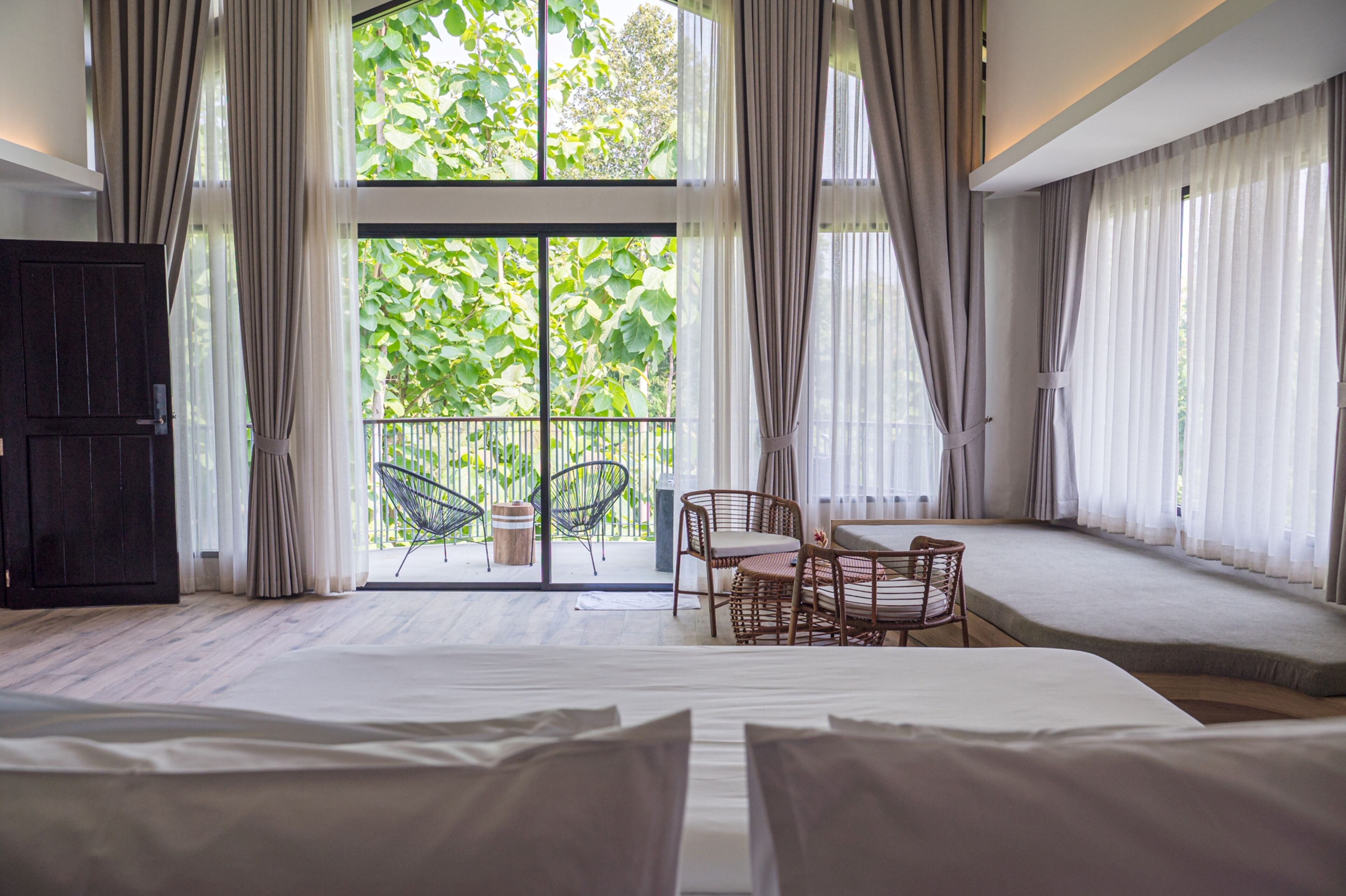 Quarto com uma cama grande, uma mesinha com cadeiras e vista para o verde através de janelas amplas.