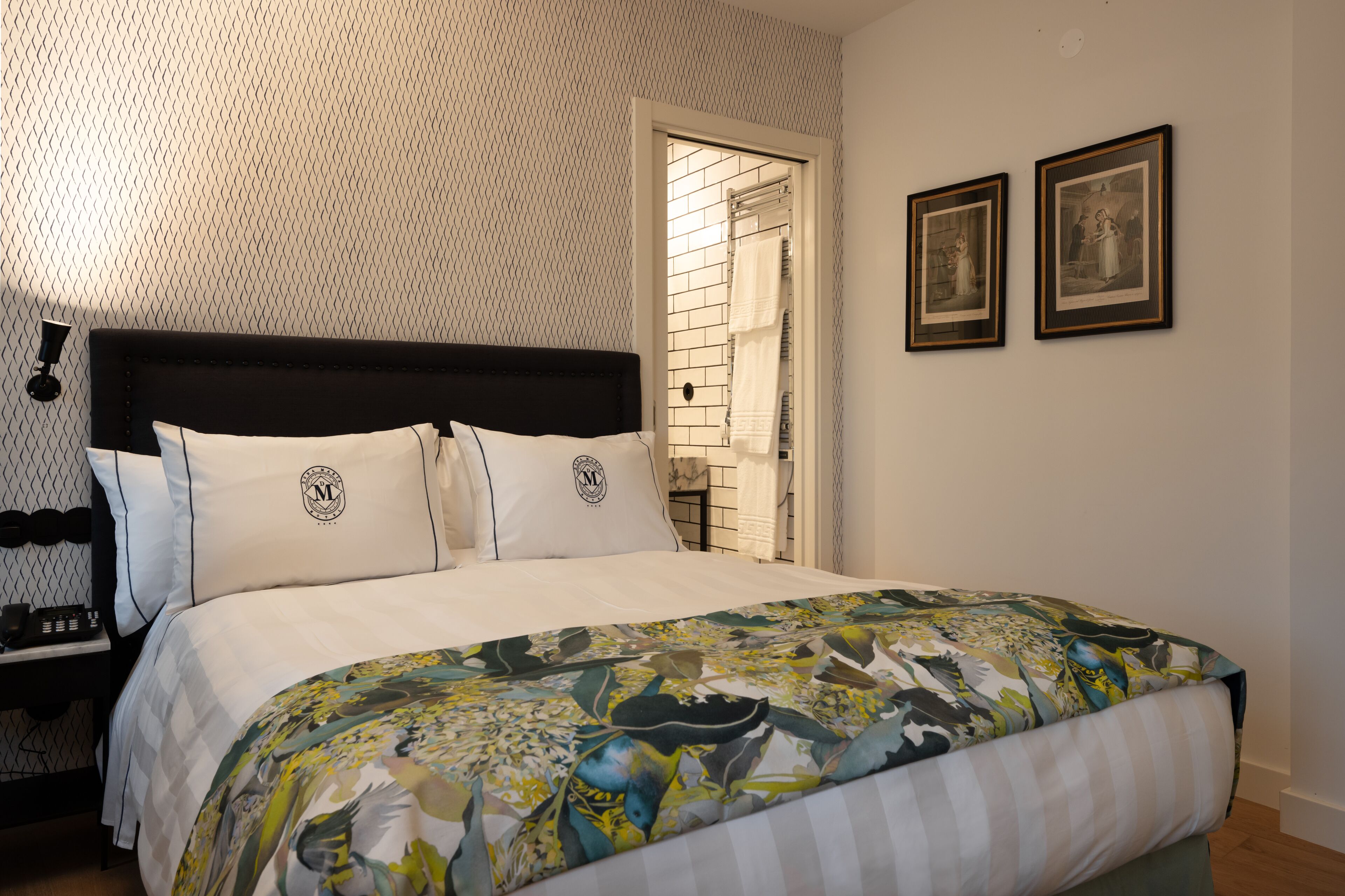 Uma cama bem arrumada com um edredom floral, uma cabeceira com um logotipo e uma mesa de cabeceira com um telefone.