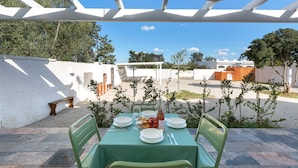 Uma área de refeições ao ar livre com uma mesa posta para dois, cercada por vegetação e uma parede branca.