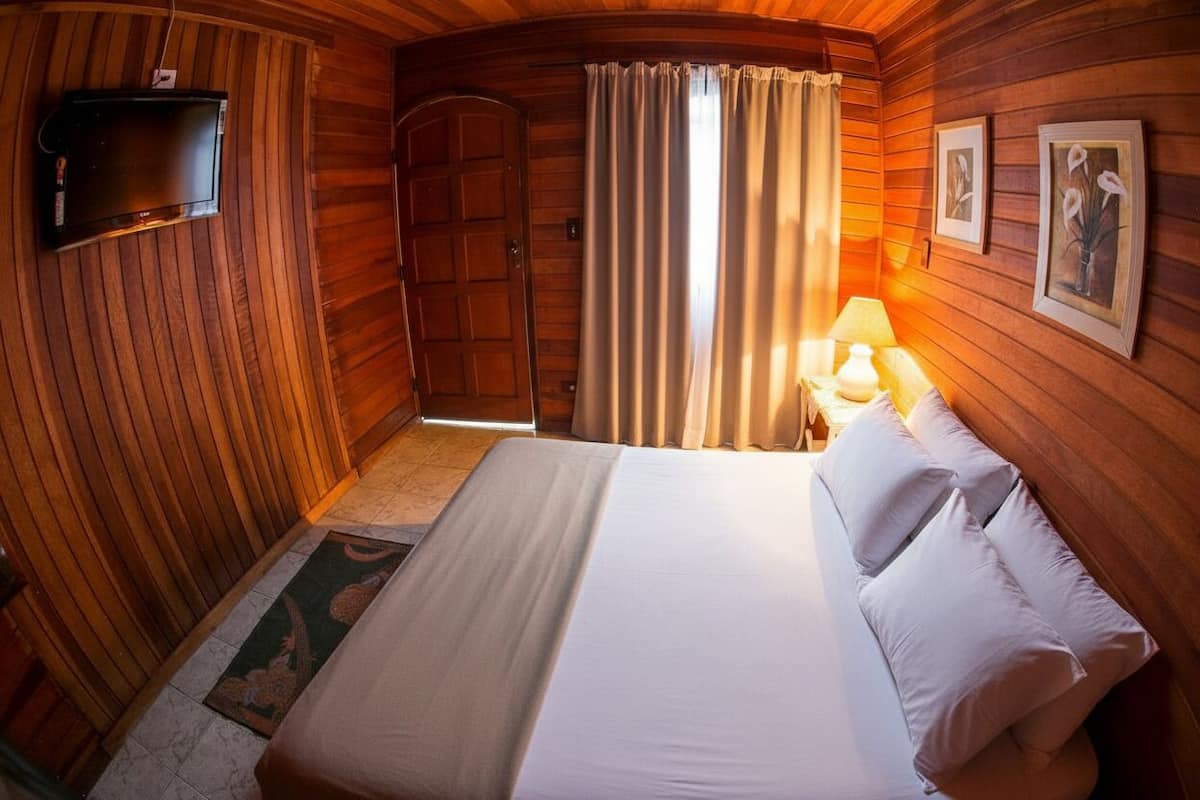 Quarto de madeira com cama, televisão, cortinas e quadros nas paredes.