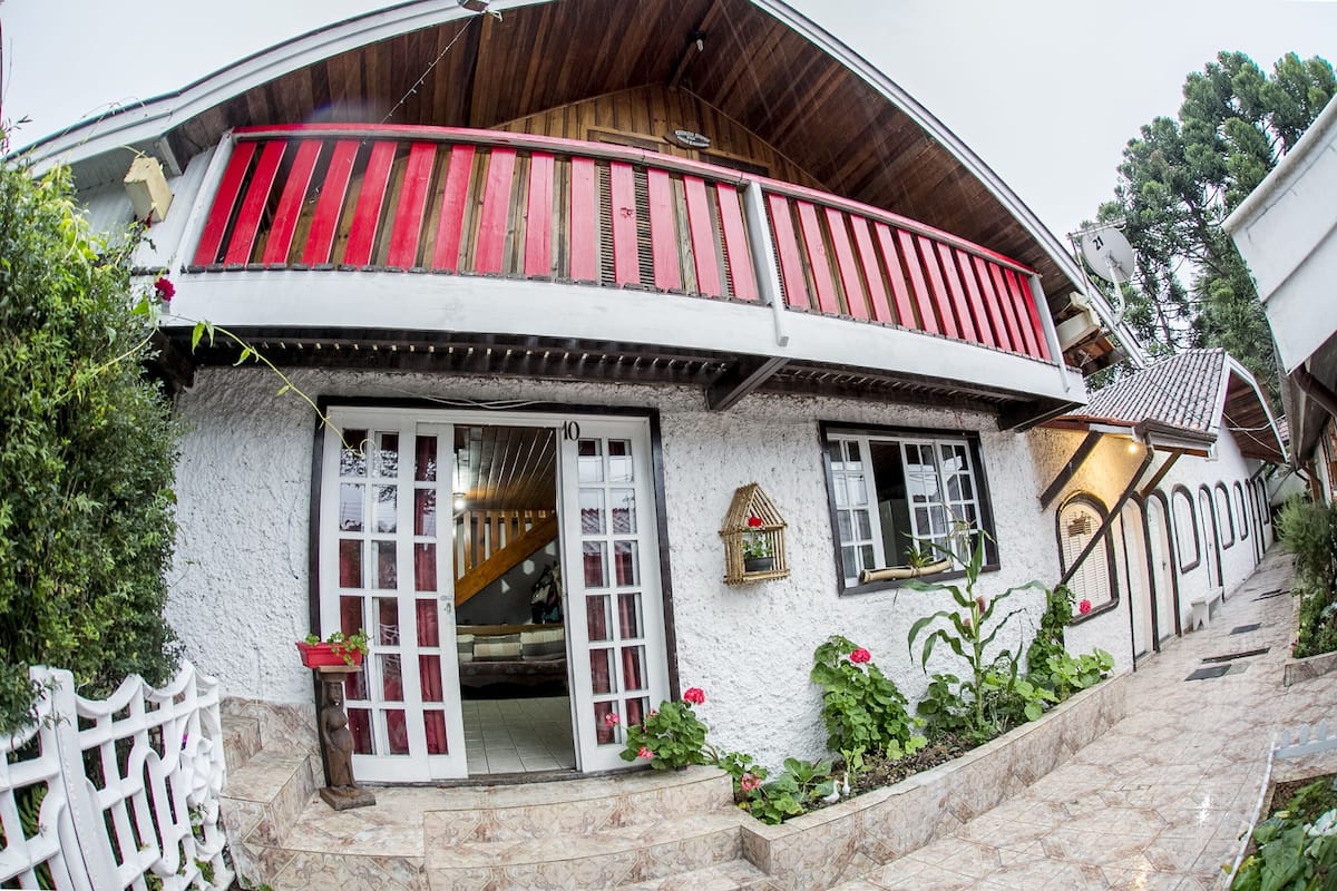Uma casa tradicional com varanda de madeira, fachada branca e um pequeno jardim com flores vermelhas.