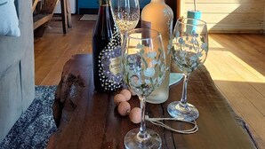 Interior acolhedor em madeira, com uma mesa de jantar posta com garrafas de vinho, copos e objetos decorativos. Uma escada de madeira e uma área de cozinha são visíveis ao fundo.