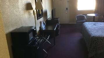 Quarto de hotel com cama, escrivaninha com computador, cadeira, mesa pequena e janela com cortinas.