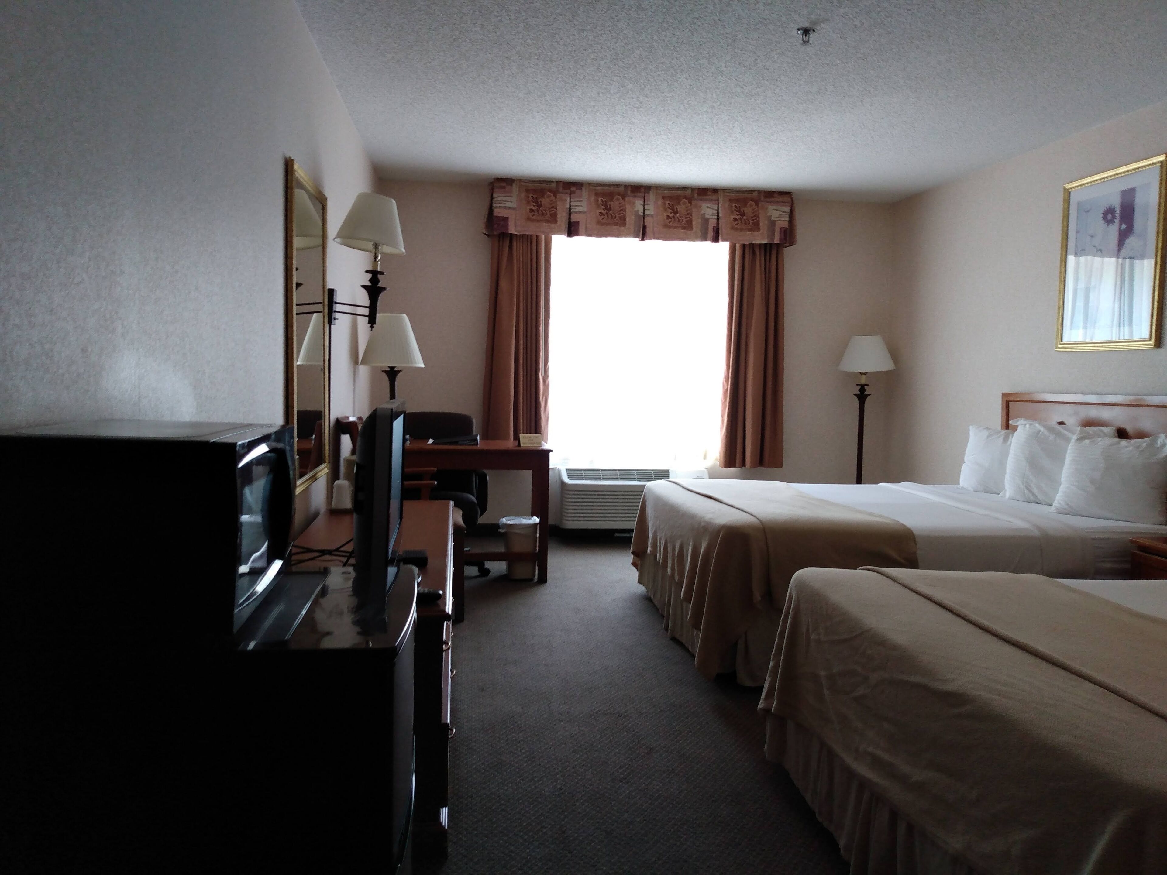 Quarto de hotel com duas camas, uma escrivaninha com computador, uma cadeira, televisão e janela com cortinas.
