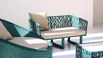 Um sofá de vime com uma almofada e uma poltrona combinando, ambos com almofada bege, em um quarto iluminado pelo sol e com piso de azulejos.