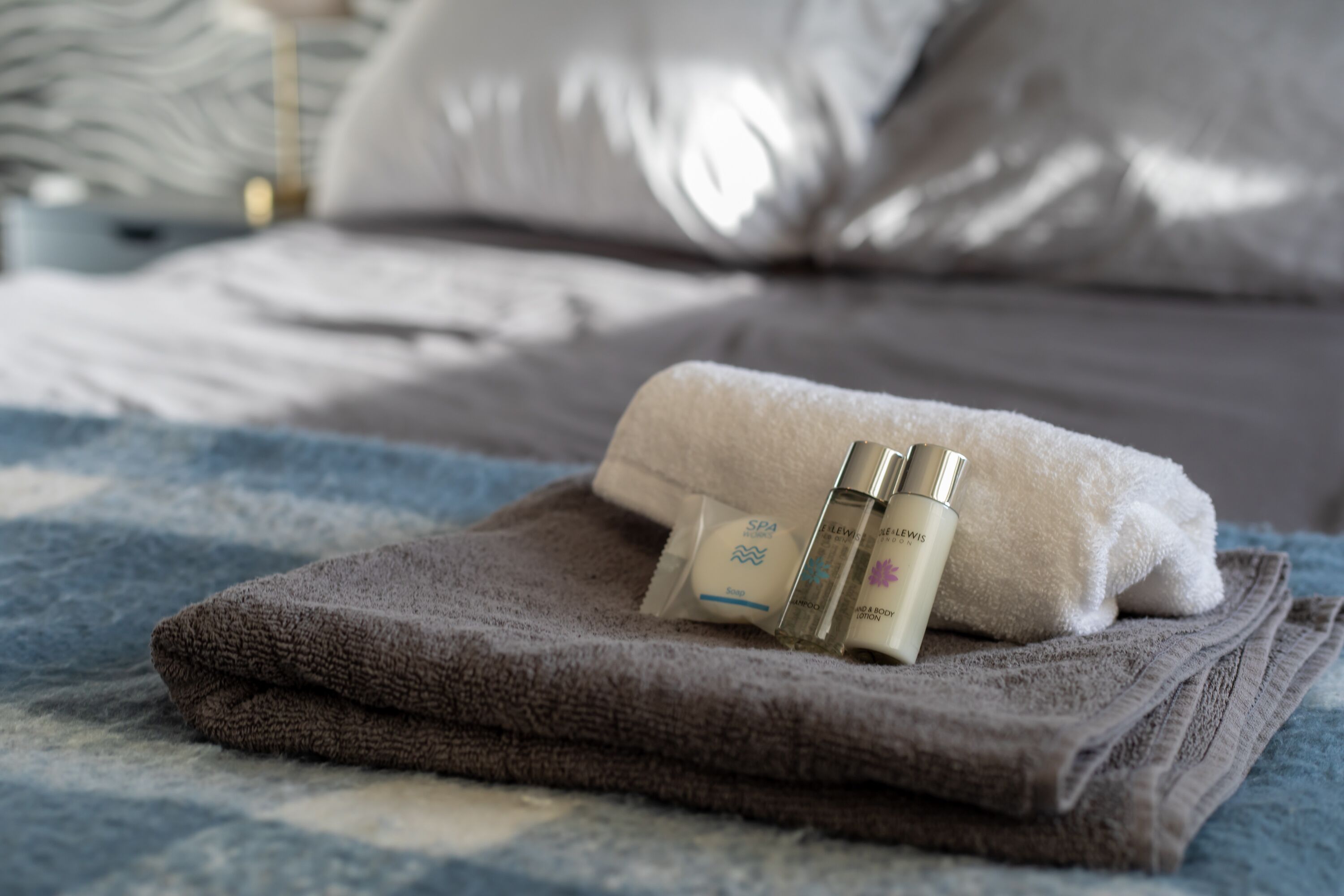 Toalha dobrada e duas embalagens de loção sobre a cama, com colcha estampada em azul e branco.