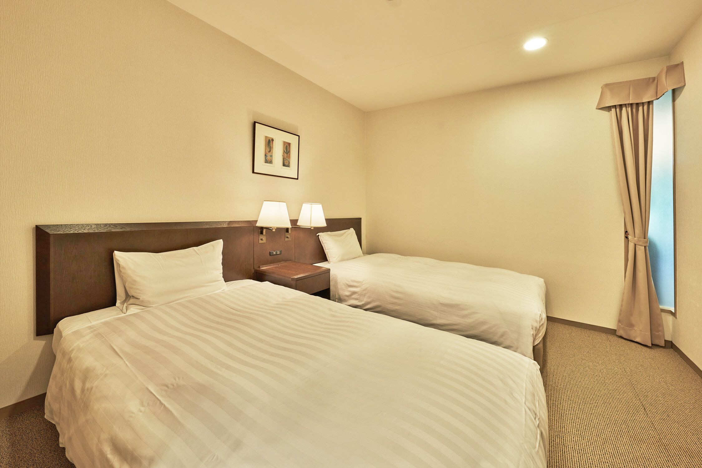 Quarto de hotel com duas camas, uma mesa de cabeceira com luminárias, um quadro na parede e uma janela com cortinas.
