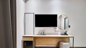 Quarto de hotel moderno com escrivaninha de madeira, televisão de tela plana, espelho redondo, secador de cabelo e lixeira.