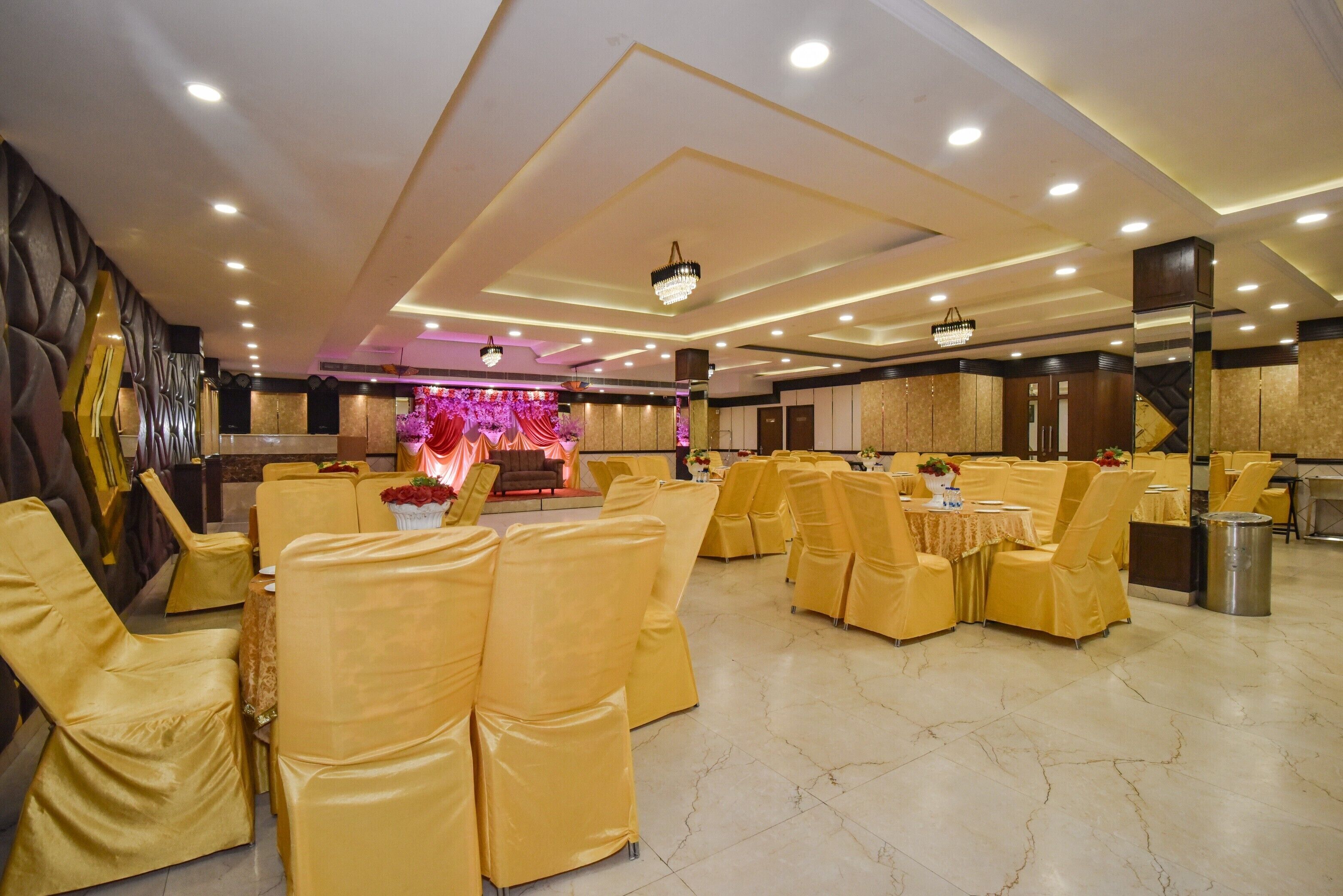 Uma sala de banquetes com mesas redondas, cadeiras amarelas e um palco com decorações florais.