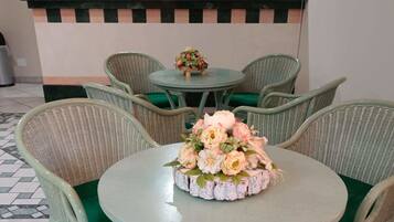 Uma sala de jantar com uma mesa redonda, cadeiras estofadas verdes e um arranjo floral. Ao fundo, um bar com diversas garrafas e uma televisão.